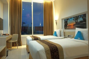 Quarto de hotel com duas camas, uma escrivaninha, uma cadeira e uma janela grande com cortinas.