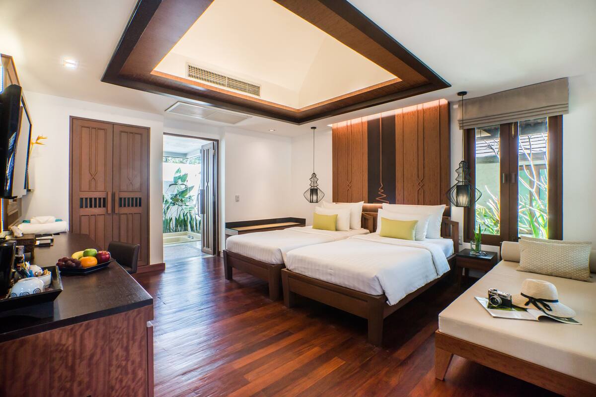 Quarto de hotel com duas camas, uma escrivaninha com fruta, uma mesa pequena e vista para o verde lá fora.
