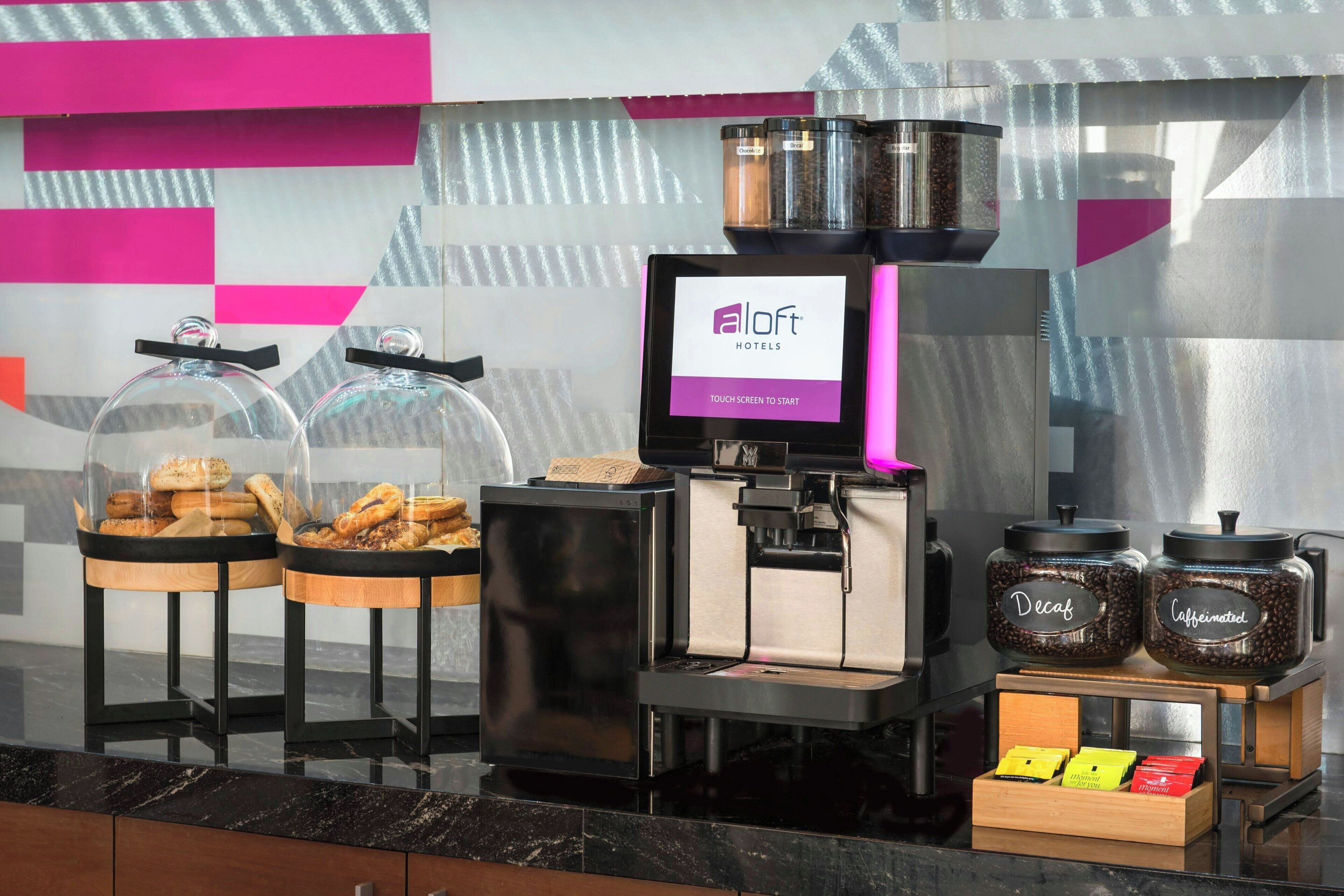 Uma estação de café com um quiosque touchscreen, dois dispensadores de café e recipientes com as etiquetas 'Decaf' e 'Cafeína'.