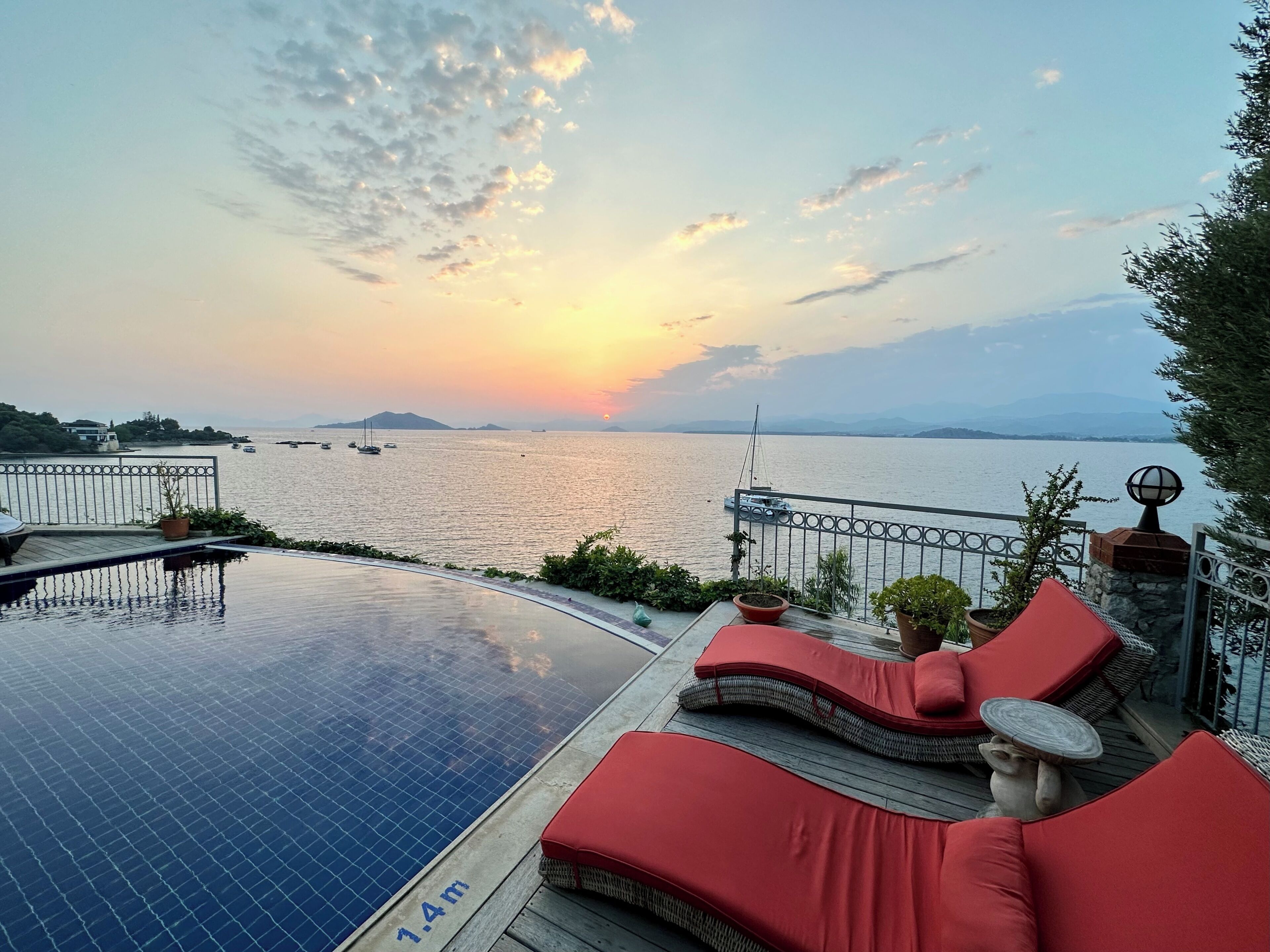 Área da piscina com vista para o mar e para o pôr do sol, com espreguiçadeiras e um vaso de planta.