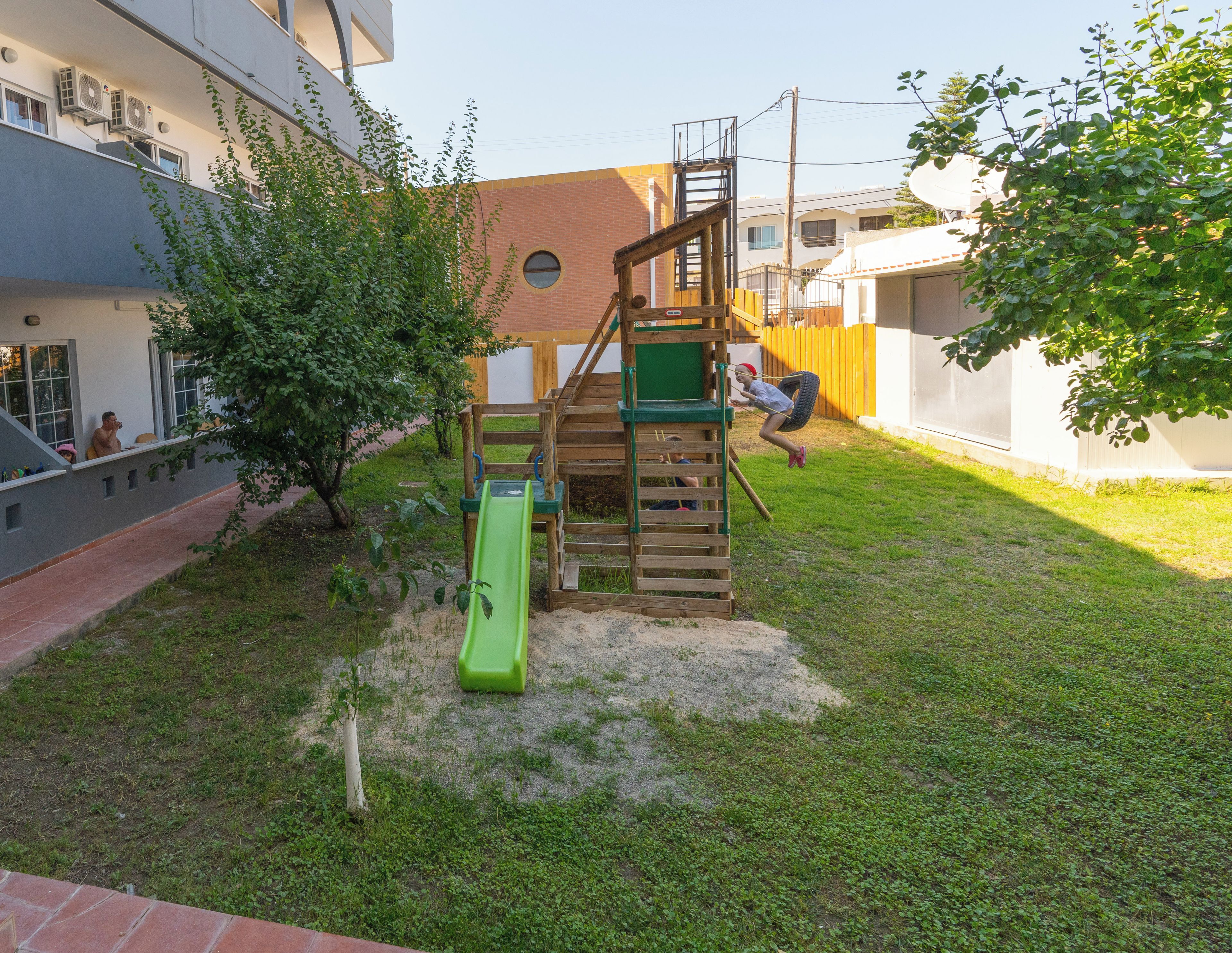 Parque infantil com escorrega e estrutura de escalada, rodeado por gramado e um edifício com janelas.