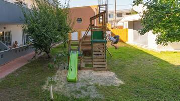 Parque infantil com escorrega e estrutura de escalada, rodeado por gramado e um edifício com janelas.