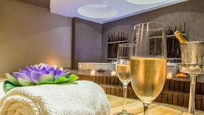 Ambiente de spa com uma toalha branca enrolada, duas taças de champanhe e uma flor roxa decorativa.