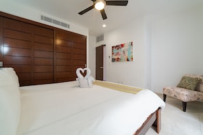 Quarto com uma cama grande, um ventilador de teto, um guarda-roupa de madeira, uma obra de arte na parede e uma poltrona estampada.