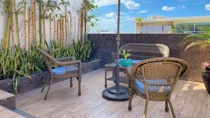Um pátio com mobiliário de vime, uma mesa e um guarda-sol. Há plantas e uma parede com bambu.