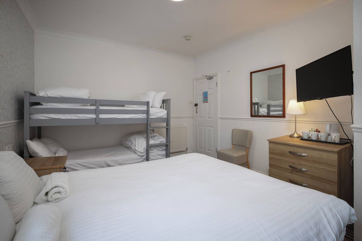 Quarto de hotel com beliche, uma cama de solteiro, penteadeira de madeira, espelho, televisão e porta.