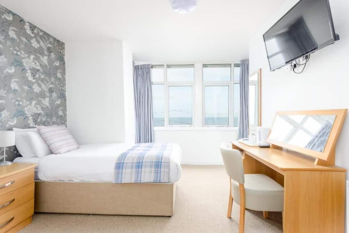 Quarto de hotel com cama, escrivaninha, cadeira, televisão e janela com cortinas.