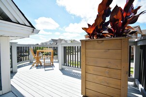 Um deck de madeira com guarda-corpo branco, um vaso de madeira com folhas grandes e um conjunto de mesa e cadeira de madeira.