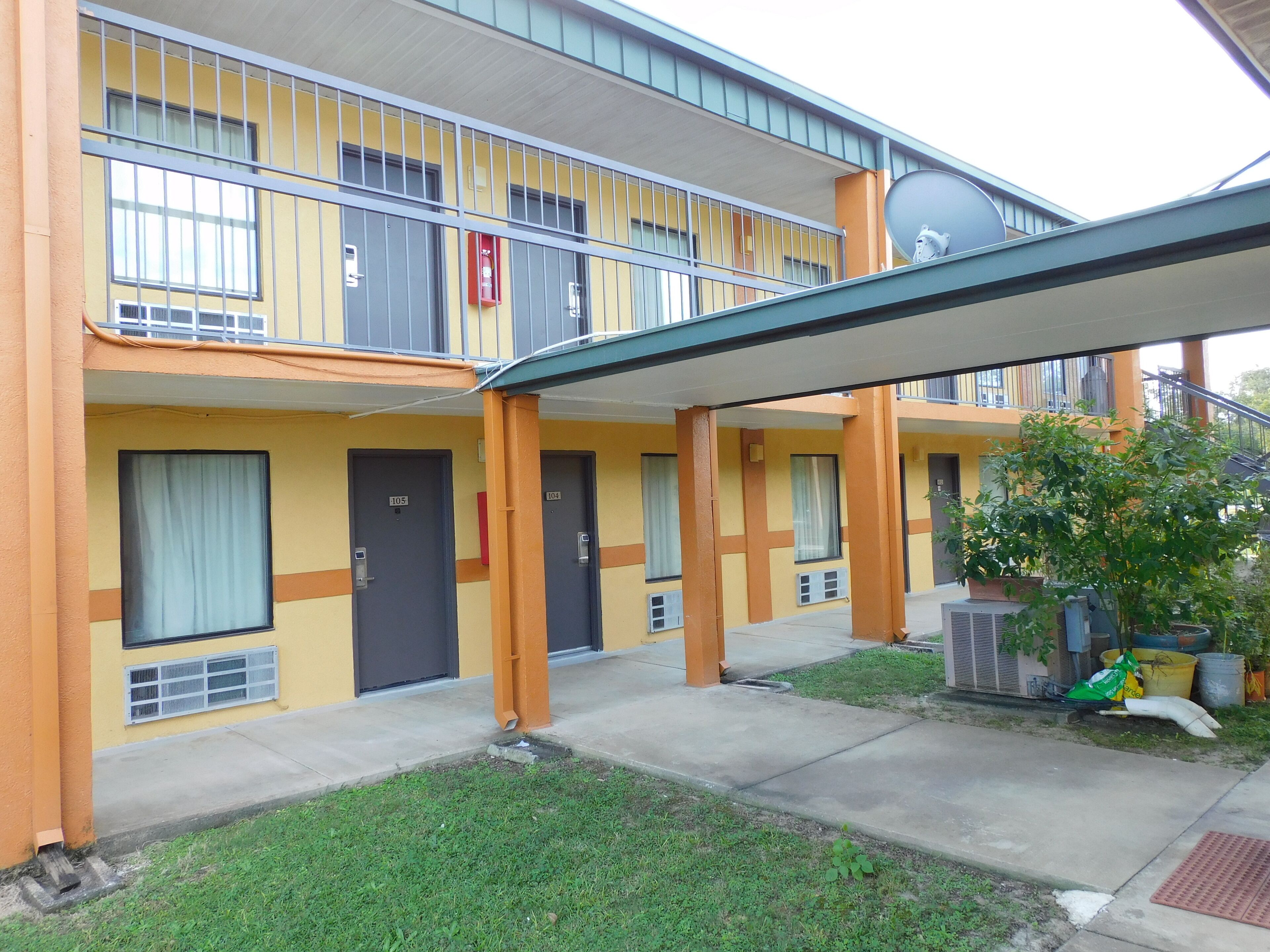 Um motel de dois andares com portas numeradas, área de estacionamento coberta e um pequeno jardim.