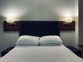 Cama bem arrumada com lençóis brancos e duas luminárias fixadas na parede, uma de cada lado.