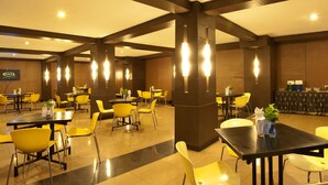 Uma área de refeições de hotel com cadeiras e mesas amarelas, um pilar grande e uma tela de televisão na parede.