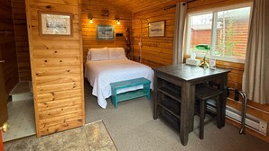 Interior de uma cabana de madeira com uma cama, uma mesa pequena, uma janela e um letreiro que diz 'WELCOME'.