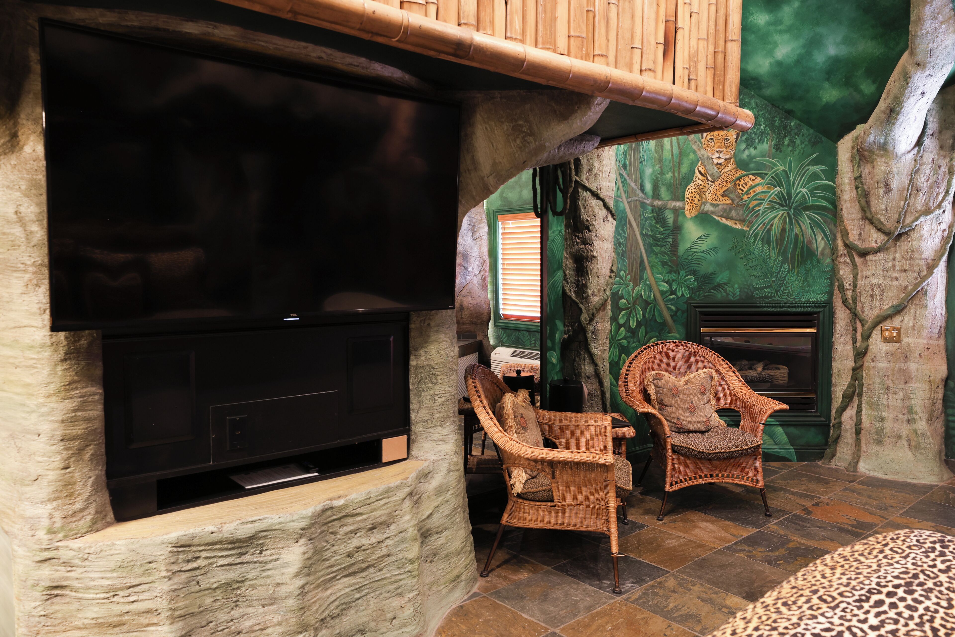 Quarto com uma televisão de tela plana grande presa a uma parede de pedra, cadeiras de vime e uma lareira com um mural verdejante de uma cena de selva.
