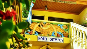 Um hotel chamado "HOTEL OLYMPOS", com fachada colorida e varanda com uma bandeira.