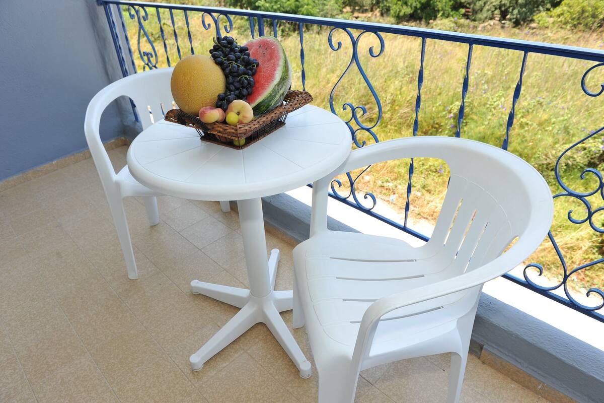 Uma varanda com uma mesa e cadeiras de plástico branco, uma cesta de vime para frutas e um corrimão de metal azul.