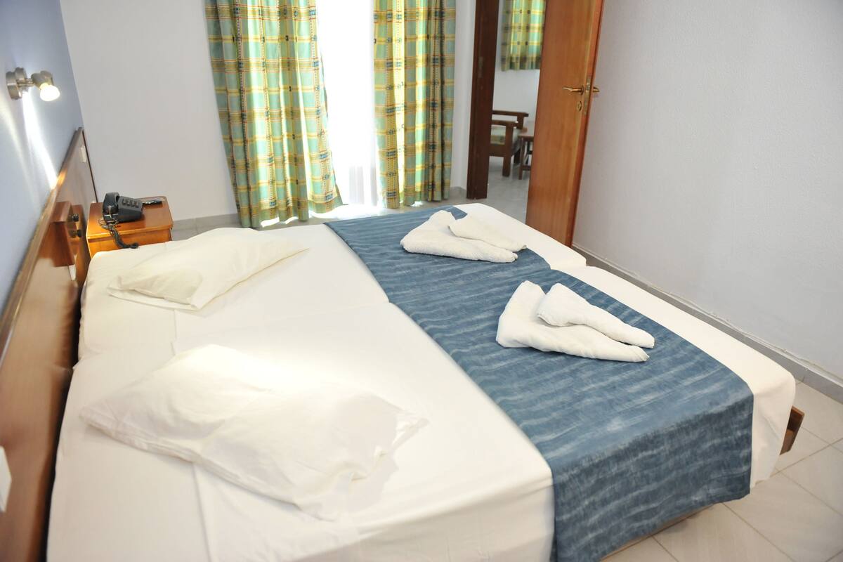 Cama bem arrumada com lençóis brancos e azuis, telefone ao lado da cama e um espelho refletindo o quarto.