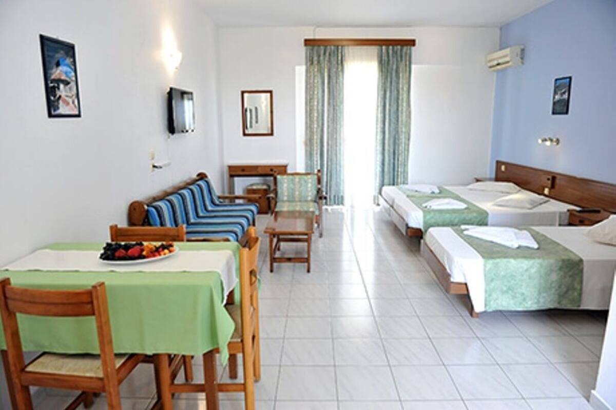 Quarto de hotel com área de jantar, duas camas de solteiro, um sofá, uma mesa com um prato de frutas e uma janela com cortinas.