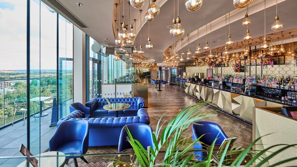 Uma área de bar com teto alto e luminárias pendentes, um espaço de estar com cadeiras azuis e vista através de grandes janelas.
