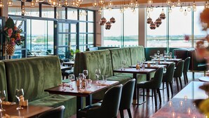 Restaurante com cabines estofadas em verde, mesas de madeira e luminárias pendentes.