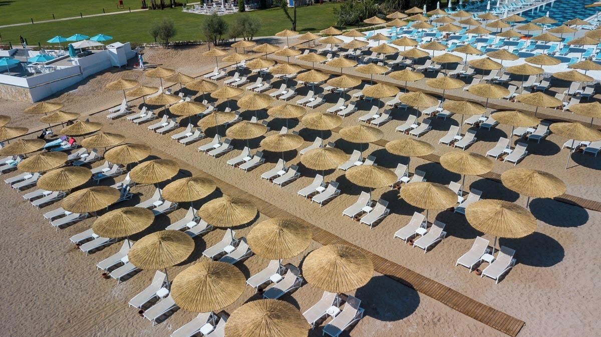 Vista aérea de um resort à beira-mar com fileiras de espreguiçadeiras e guarda-sóis de palha, área de piscina e um gramado.