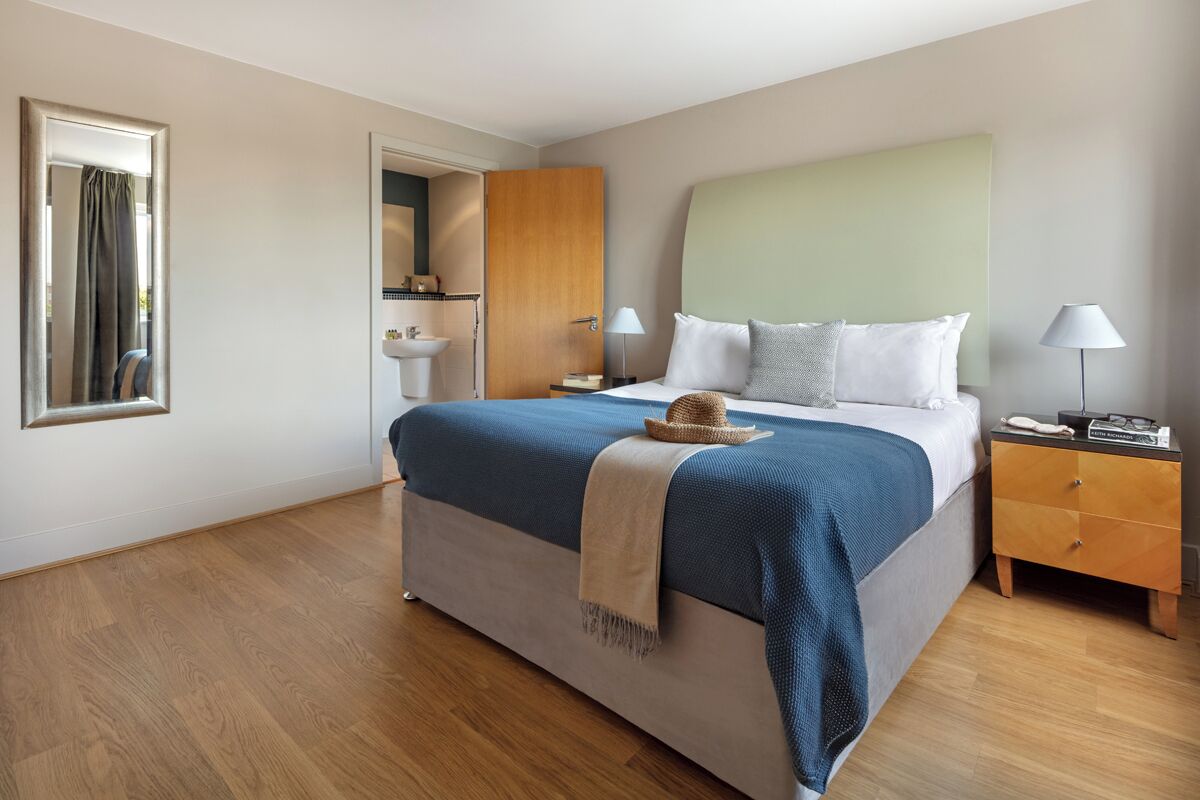 Quarto de hotel moderno com uma cama grande, uma mesa de cabeceira, um guarda-roupa e um banheiro visível através de uma porta aberta.