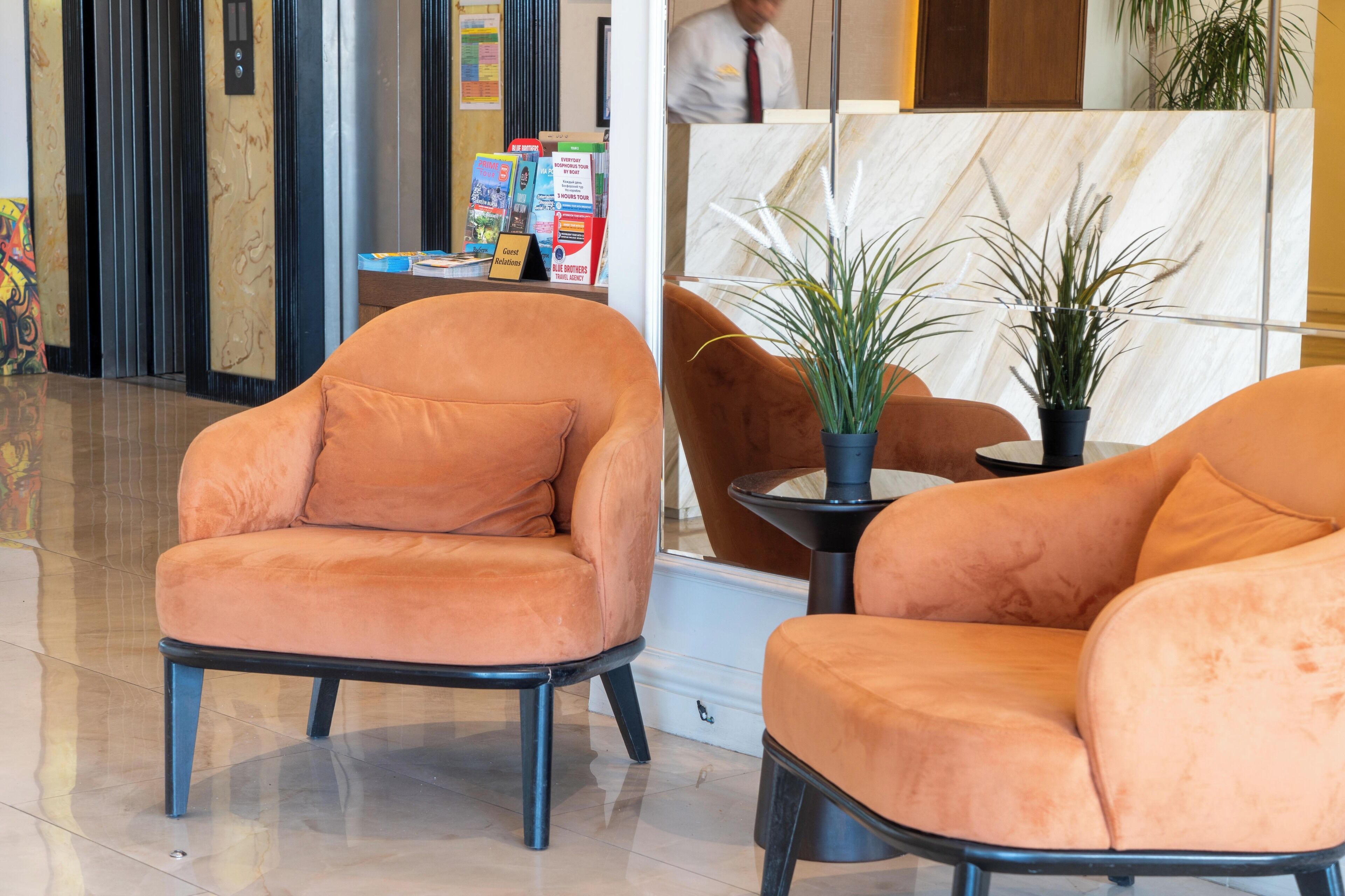 Um hall de hotel com balcão de recepção, área de estar com cadeiras laranja e uma planta em vaso.