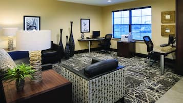Quarto de hotel com sofá, escrivaninha com computador, impressora e janela com vista para a cidade.