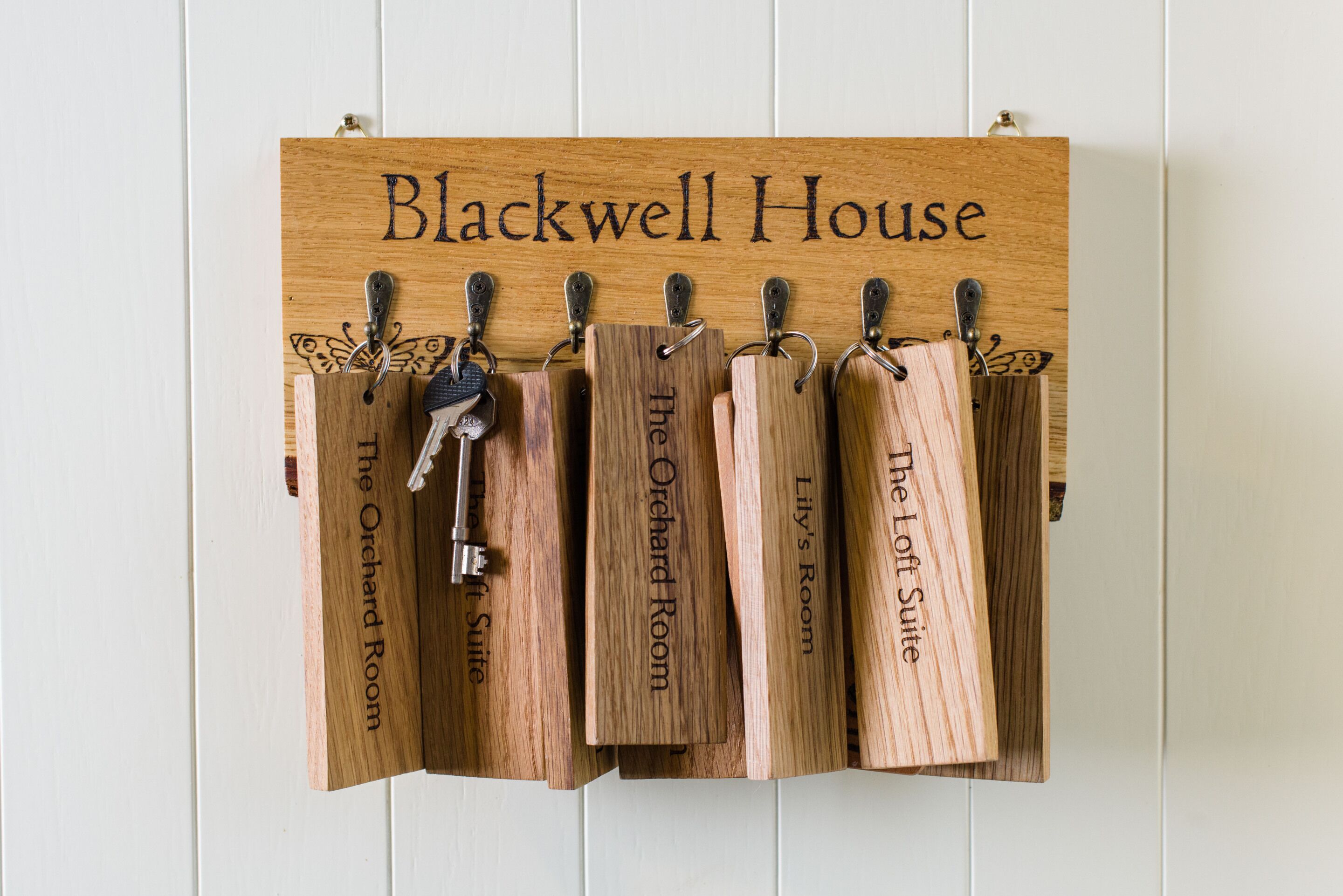 Uma placa de madeira com o nome 'Blackwell House' e um conjunto de ganchos de madeira para chaves abaixo, cada um identificado com nomes de quartos como 'The Orchard Room' e 'The Loft Suite'.