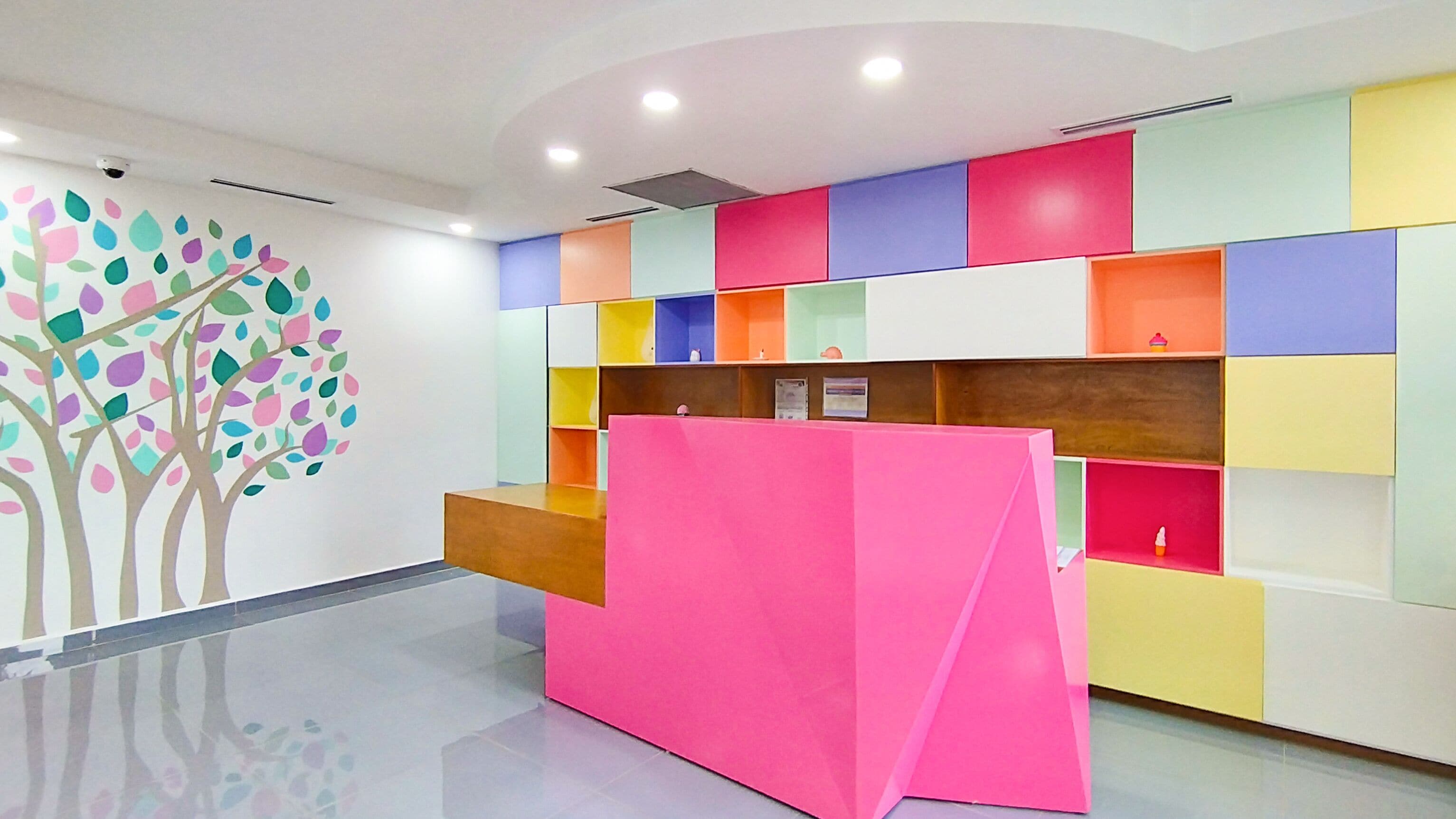 Uma área de recepção luminosa e colorida, com design moderno, apresentando uma divisória rosa grande e uma parede com um mural de uma árvore colorida.
