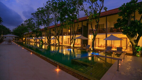 Um hotel com piscina, espreguiçadeiras, guarda-sóis e fachada bem iluminada ao anoitecer.