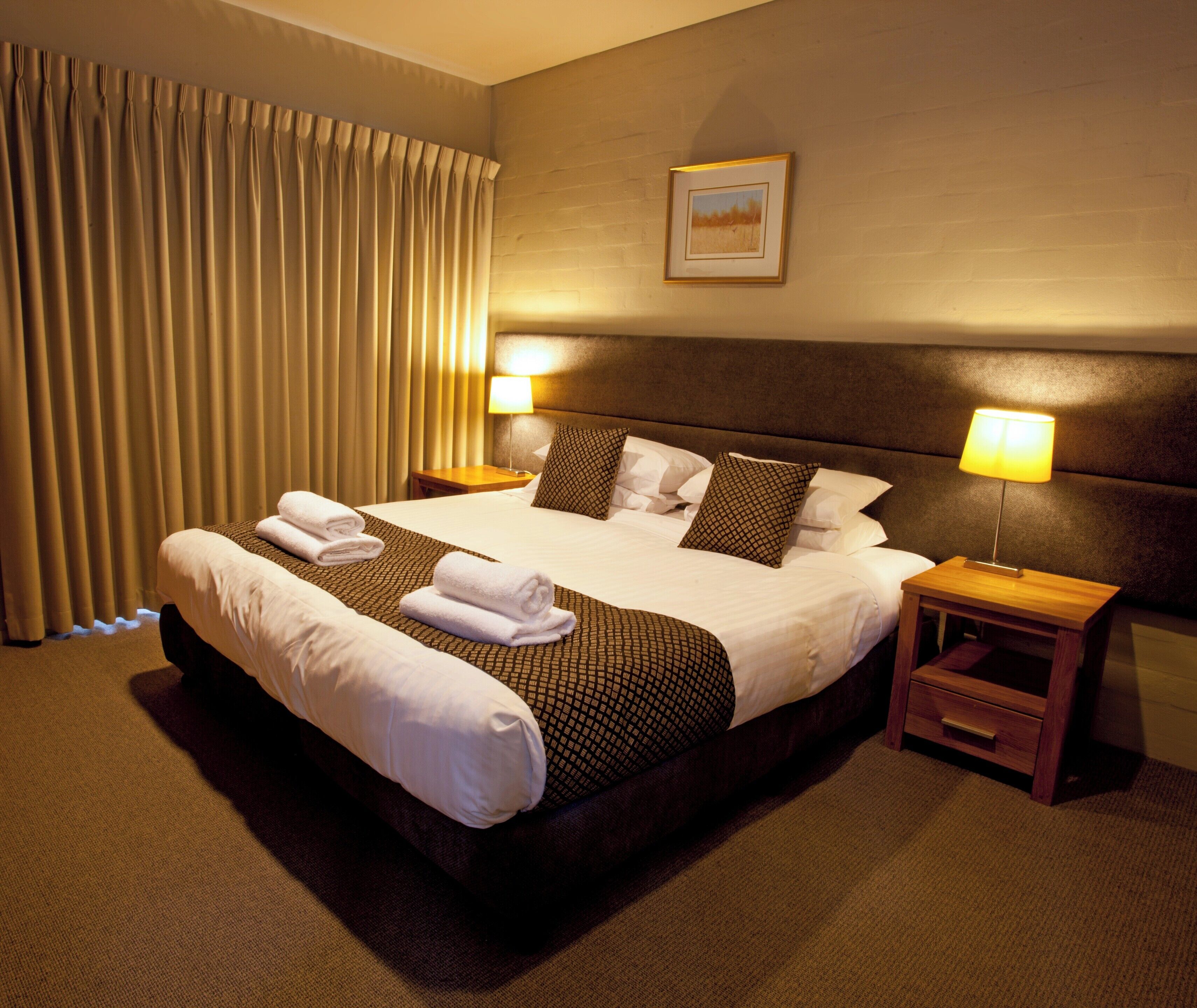 Quarto de hotel com uma cama grande, duas luminárias de cabeceira, cabeceira, um quadro na parede e uma janela com cortinas.
