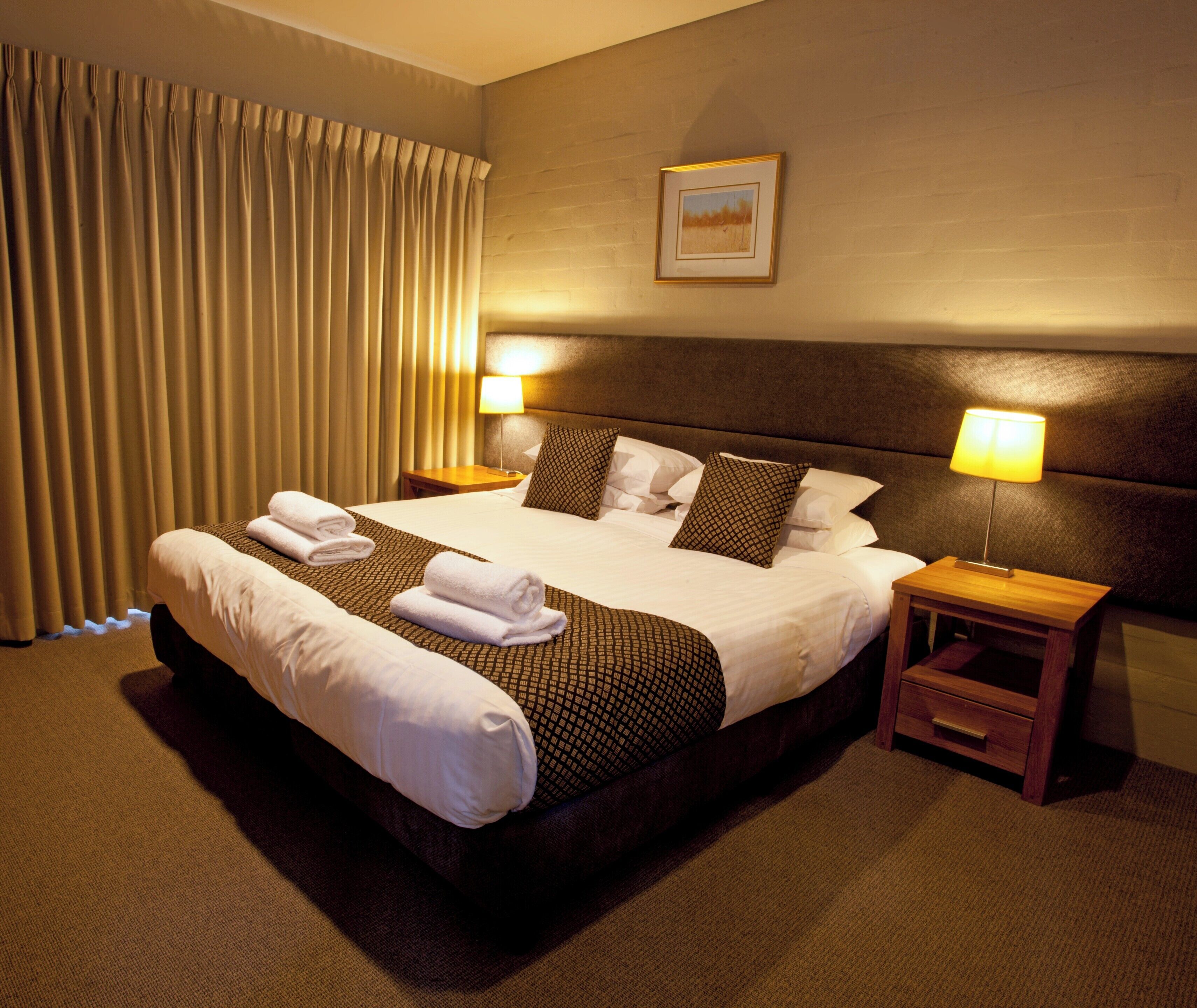 Quarto de hotel com uma cama grande, duas mesas de cabeceira, cabeceira, um quadro emoldurado na parede e uma janela com cortinas.