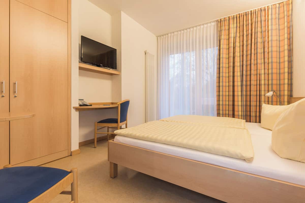 Quarto com cama, escrivaninha com cadeira, televisão na parede e janela com cortinas.