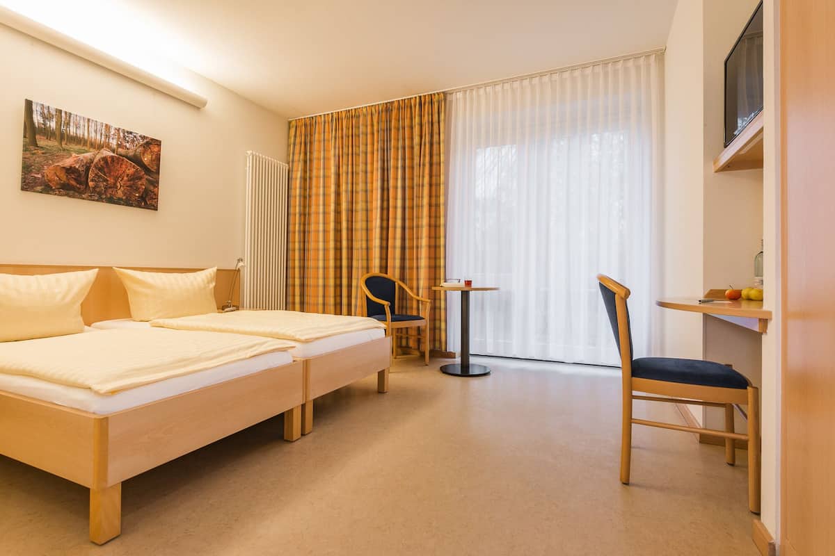 Quarto de hotel com duas camas de solteiro, uma cadeira, uma mesa pequena, um radiador e um quadro na parede.