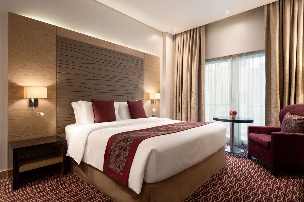 Quarto de hotel com uma cama grande, duas luminárias de cabeceira, uma mesinha com um vaso, uma poltrona vermelha e vista para o exterior através de cortinas translúcidas.