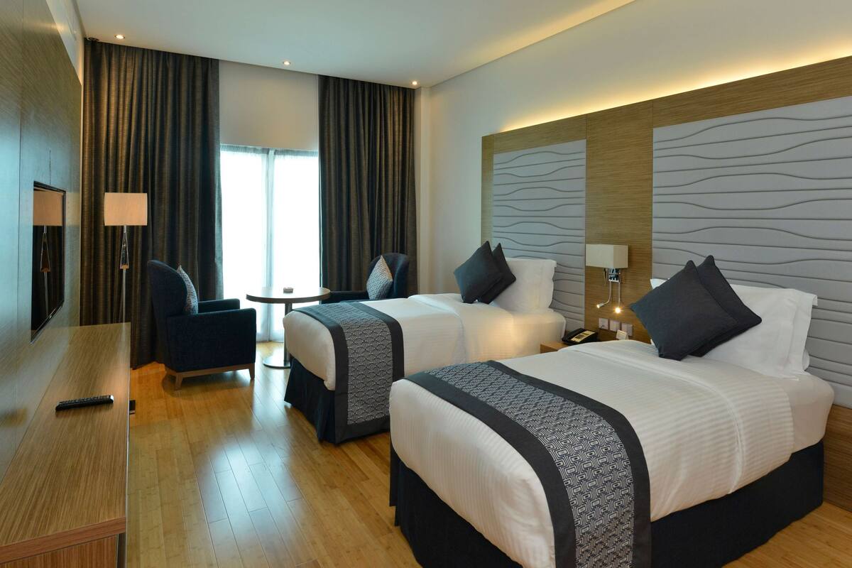 Quarto de hotel com duas camas, uma escrivaninha, uma cadeira, uma televisão e uma janela com cortinas.