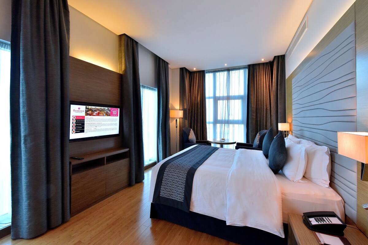 Quarto de hotel moderno com uma cama grande, uma TV em um suporte, uma escrivaninha com luminária e vista para o exterior através de janelas amplas.