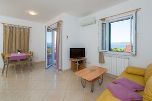 Uma sala de estar moderna com sofá, televisão, área de jantar com cadeiras e mesa, e uma janela grande com vista.