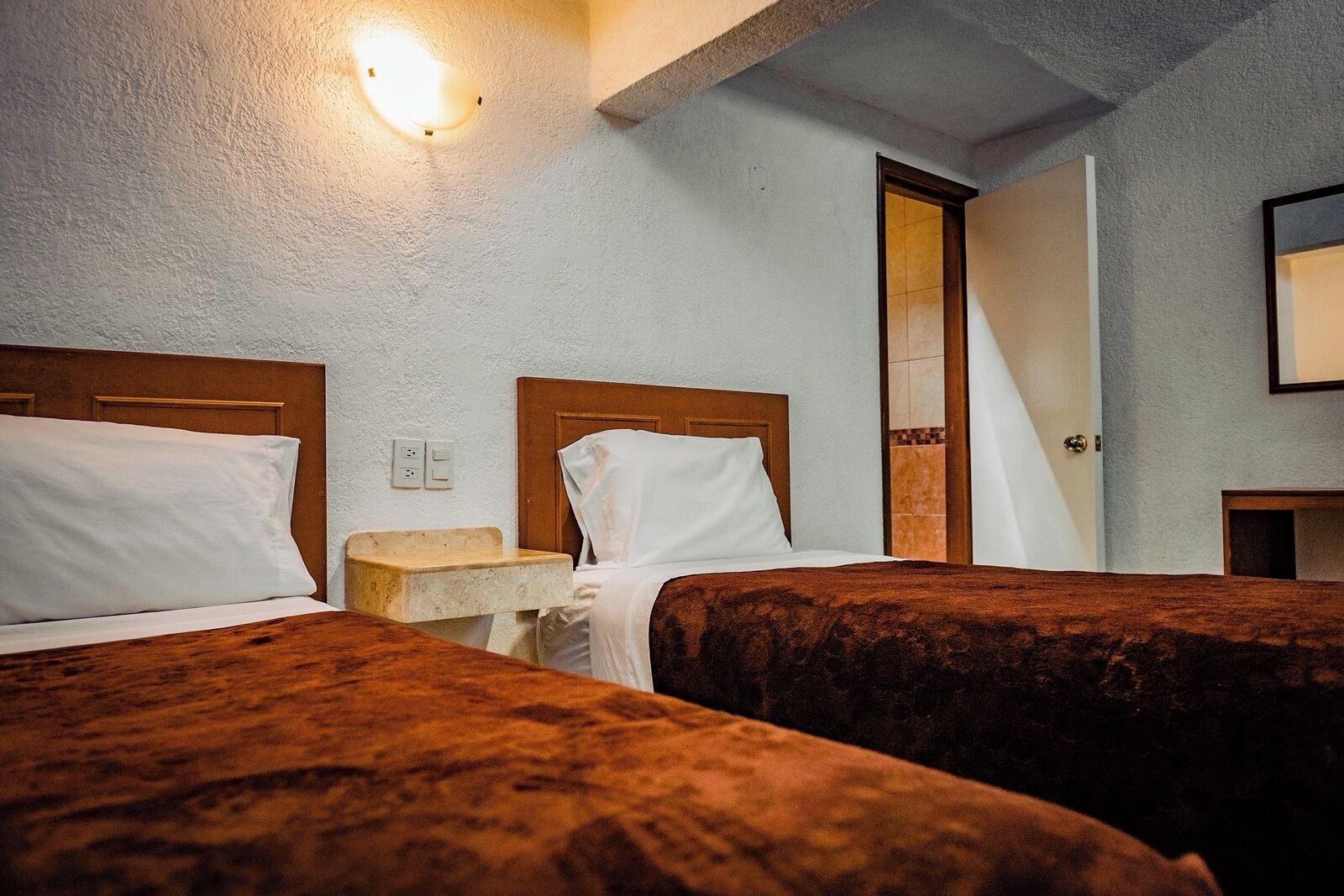 Quarto de hotel com duas camas, uma mesa de cabeceira, uma luminária na parede e uma porta que dá para outro cômodo.