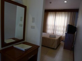 Quarto de hotel com escrivaninha de madeira, espelho grande, cama com roupa de cama listrada, televisão em suporte e janela com cortinas.