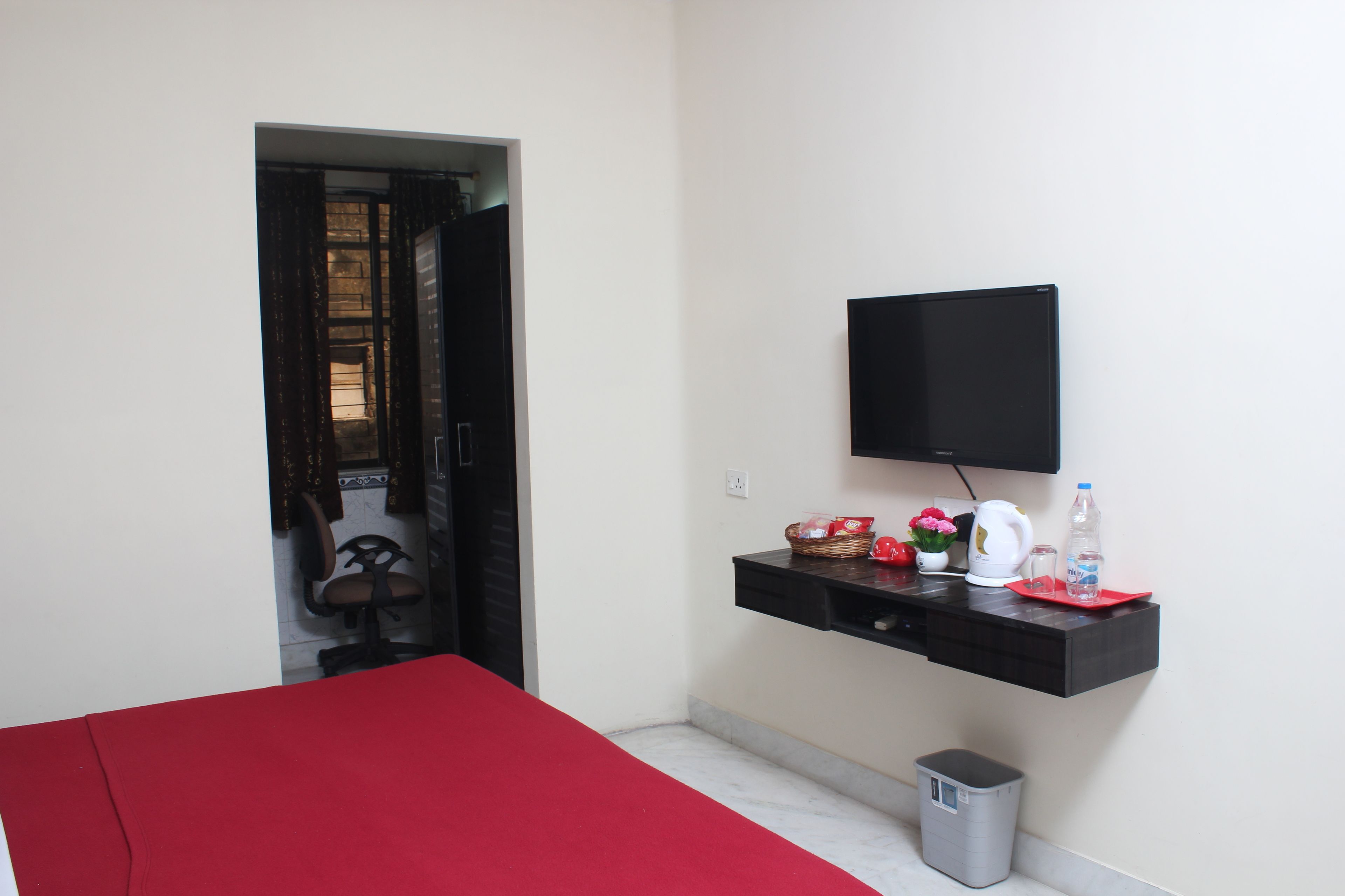 Quarto com cama vermelha, televisão preta em uma estante preta, lixeira e uma pequena mesa com cesta e um vaso de flores.