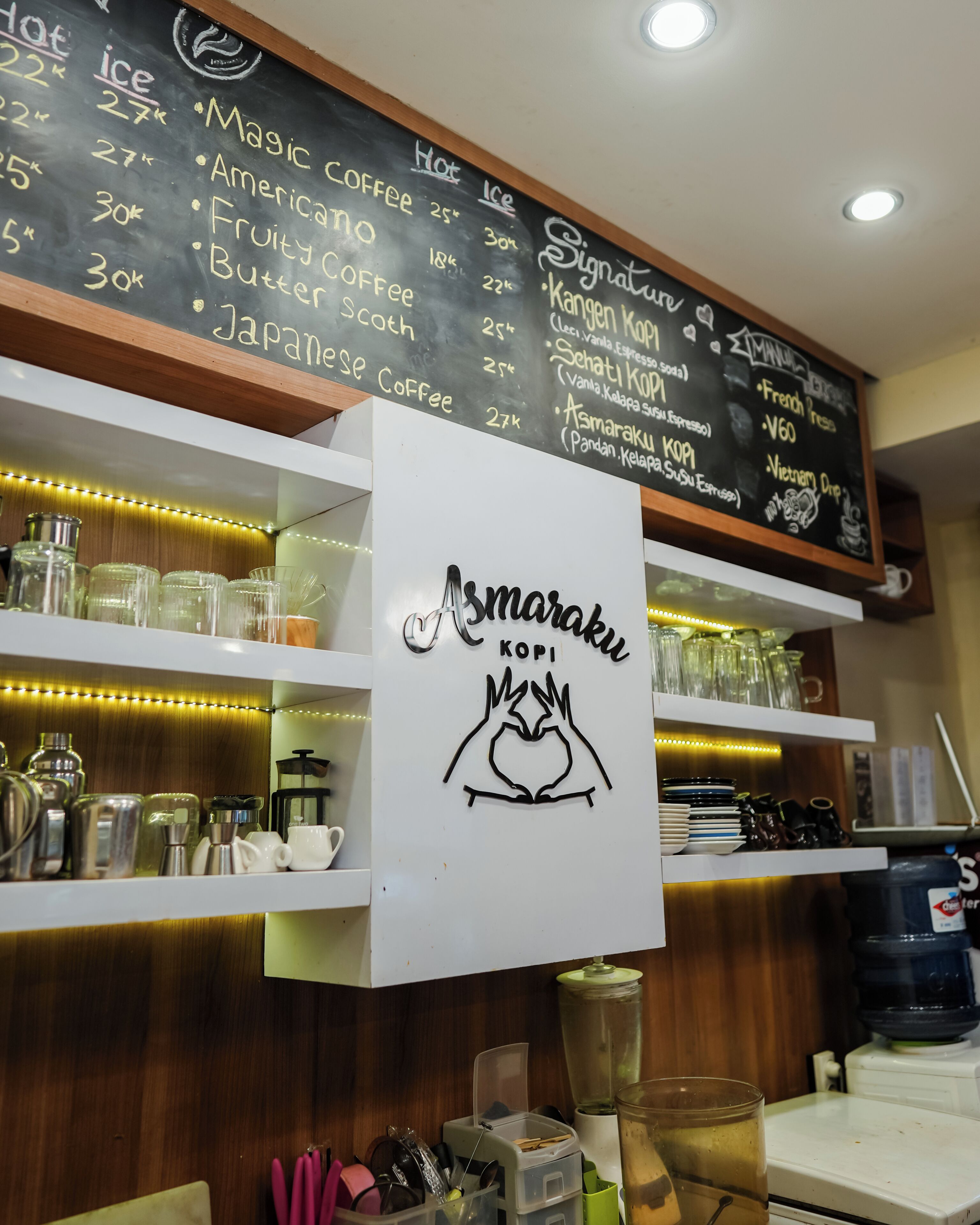 Interior de cafeteria com um quadro negro exibindo diversos tipos e temperaturas de café, um letreiro com ilustração de uma xícara de café e prateleiras com equipamentos para preparo de café.