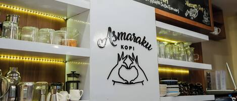 Interior de cafeteria com um quadro negro exibindo diversos tipos e temperaturas de café, um letreiro com ilustração de uma xícara de café e prateleiras com equipamentos para preparo de café.
