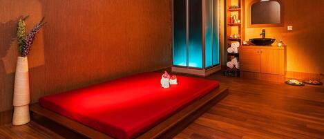 Um espaço de spa com uma mesa de massagem vermelha, um armário de madeira com produtos de higiene pessoal e um box de chuveiro com iluminação azul.
