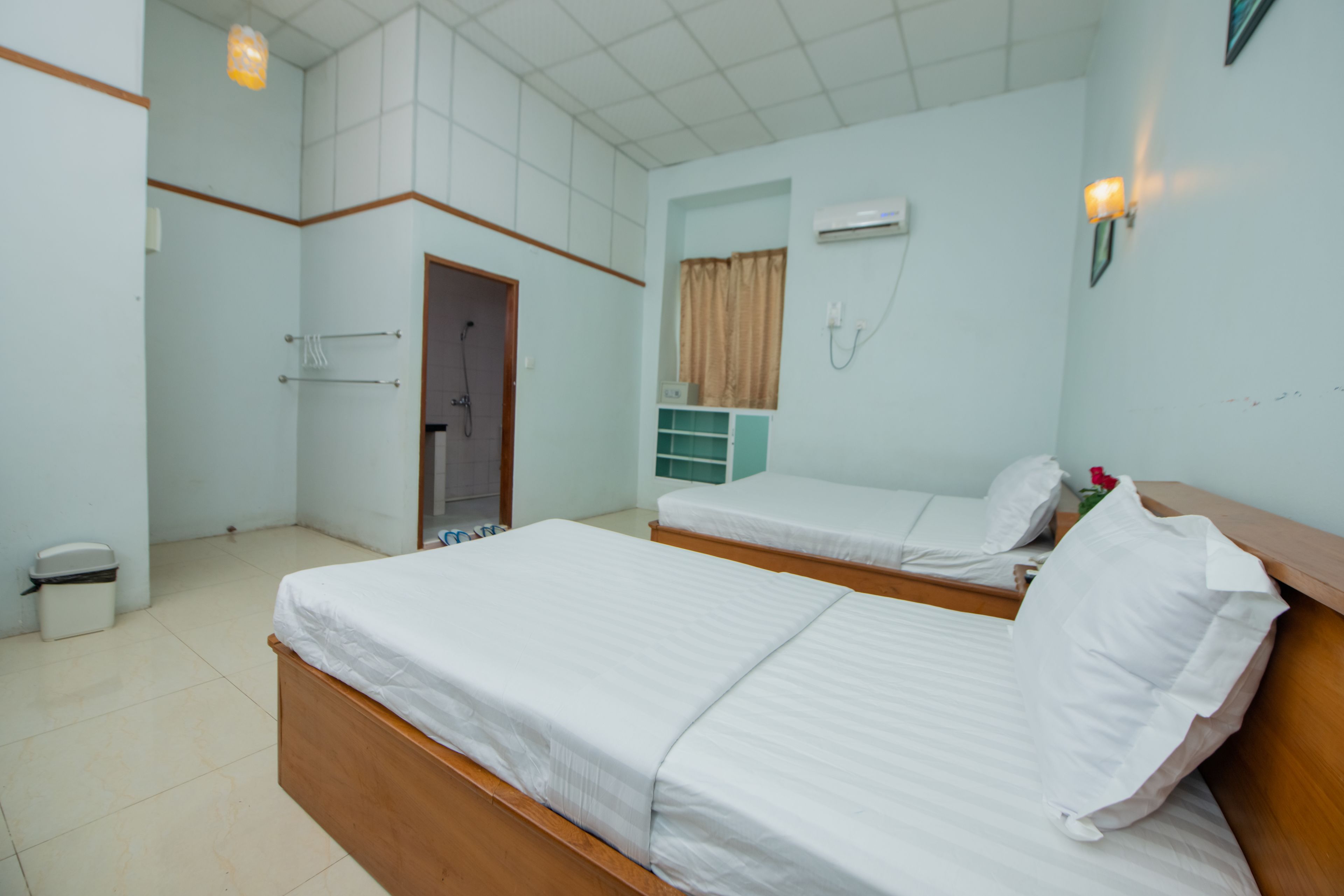 Quarto de hotel com duas camas, chuveiro, uma pequena janela com cortinas e ar condicionado fixado na parede.