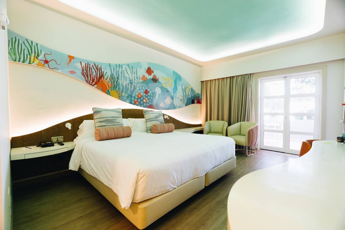 Quarto de hotel com cama, escrivaninha, cadeira, janela grande e um mural na parede.