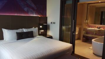 Quarto de hotel com uma cama grande, uma mesa de cabeceira com abajur, e banheiro com banheira e espelho.