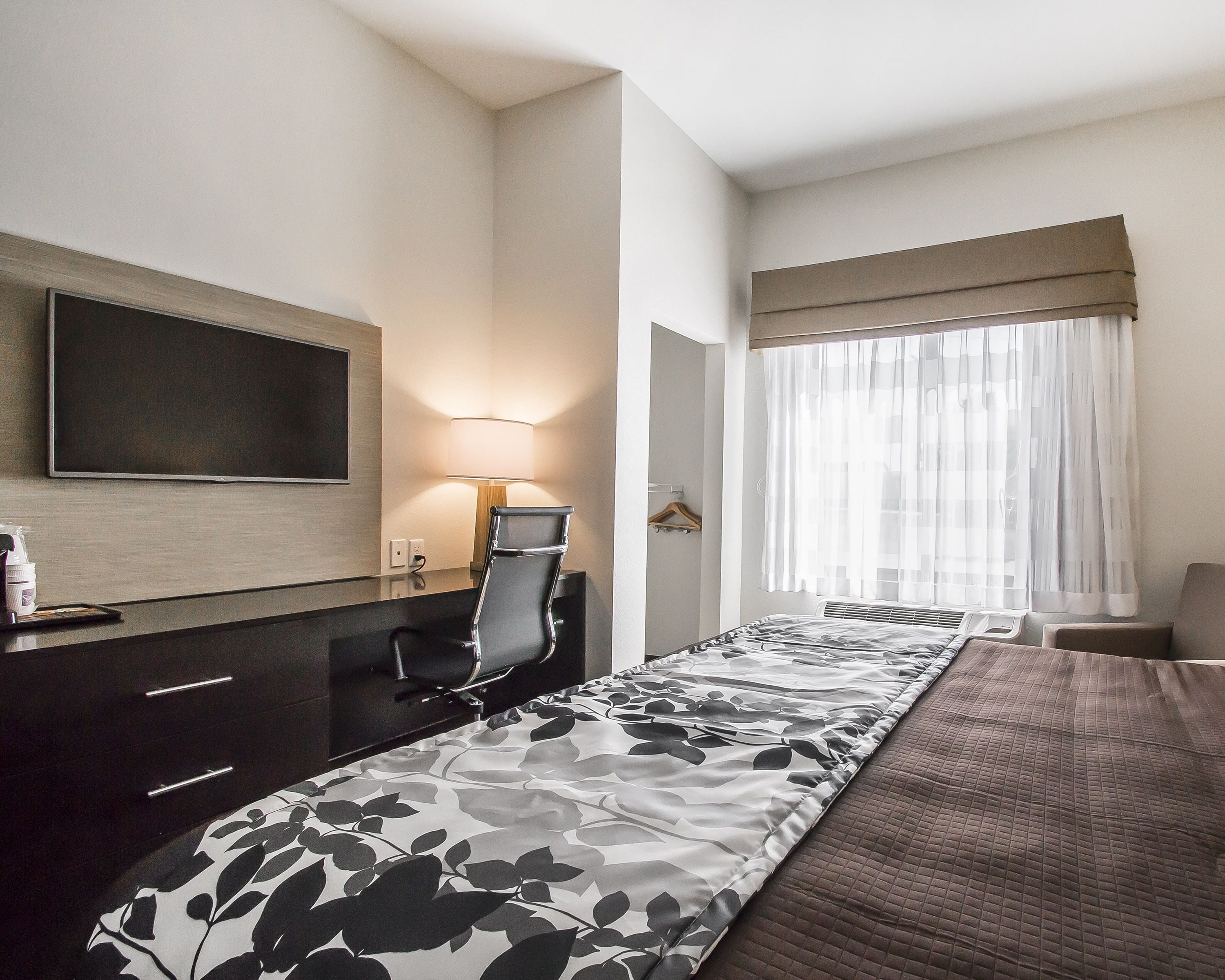 Quarto de hotel com uma cama grande, uma escrivaninha com cadeira, uma televisão de tela plana, um abajur e uma janela com cortinas finas.