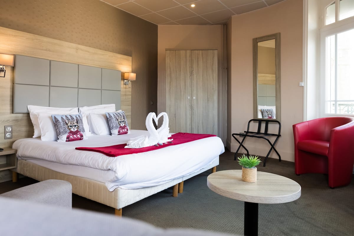 Quarto de hotel com uma cama grande, uma mesa redonda, uma cadeira vermelha e uma pequena planta.