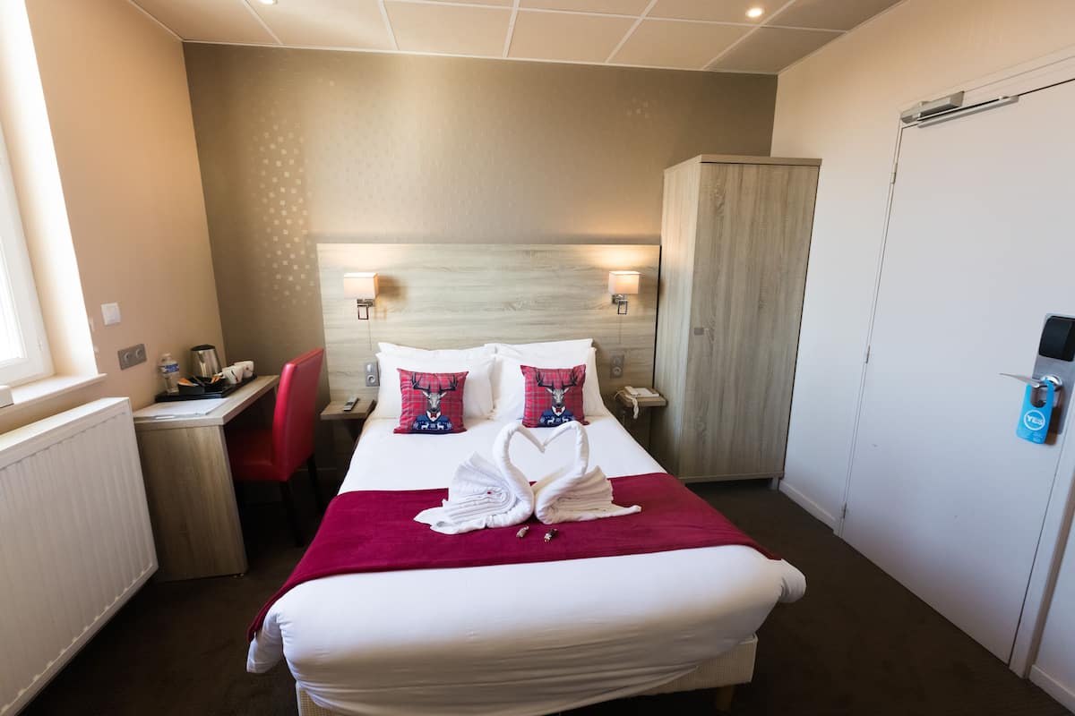Quarto de hotel com cama, escrivaninha, cadeira, guarda-roupa e mesa de cabeceira com luminária.
